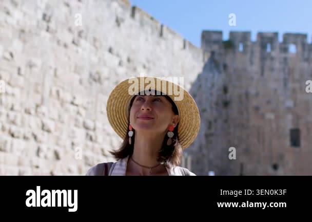 A smiling mature woman in a straw hat walks near an old stone wall in ...