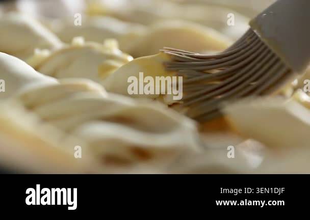 Baker applying egg wash with a brush on uncooked pastry dough pieces ...