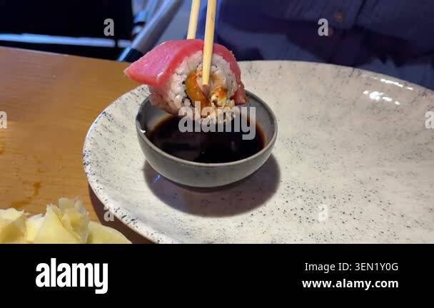 Wooden chopsticks hold a tuna sushi roll above dipping bowl as dark soy ...