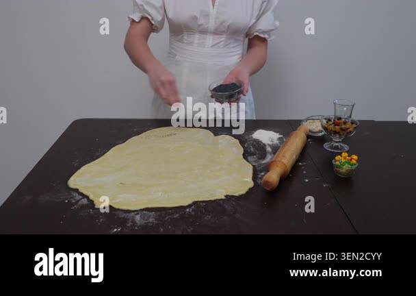 A young woman evenly scatters poppy seeds across a sheet of rolled ...