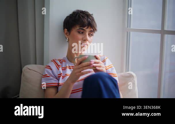 Brunette drinking hot tea sitting apartment floor looking window ...