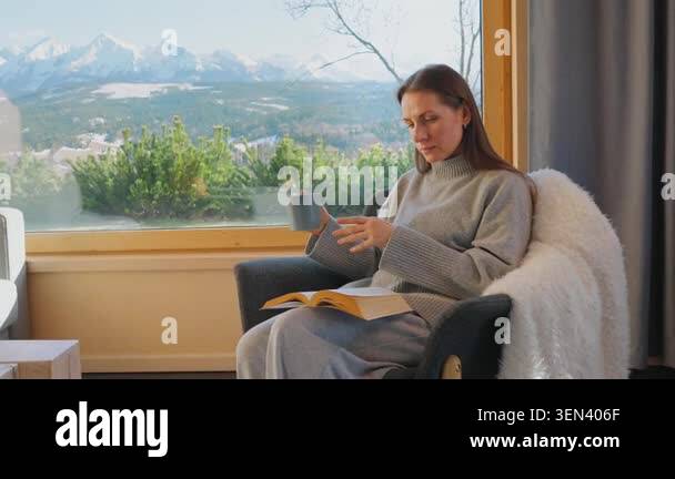 A woman reading by the large window and enjoying scenic snowy mountain ...