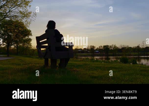 Silhouette of a woman sitting on a bench near a lake during sunset in a ...