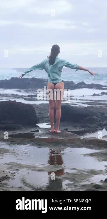 woman stretching arms on rocky shore, teal shirt and black bikini ...