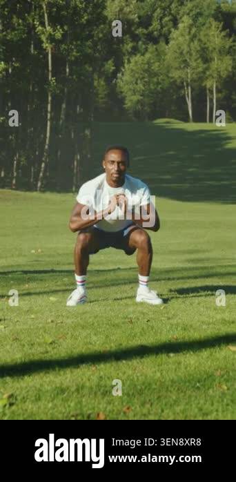 Black man performing squat workout outdoors in sunny park wearing white ...