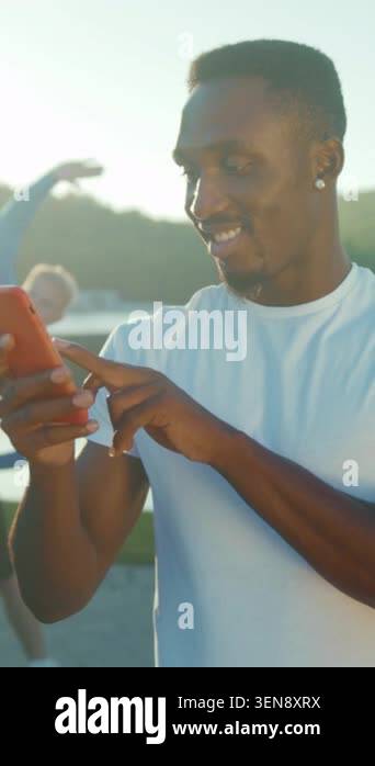 black man smiling at smartphone during lakeside sunset, tapping red ...