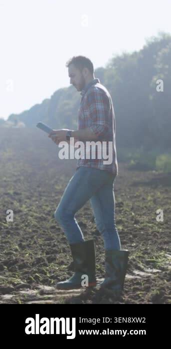 white farmer walking across ploughed field, wearing rubber boots and ...