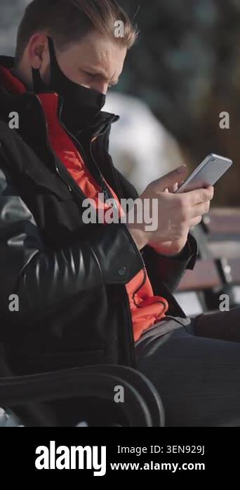 A man in a black mask and orange jacket looks at his smartphone in ...