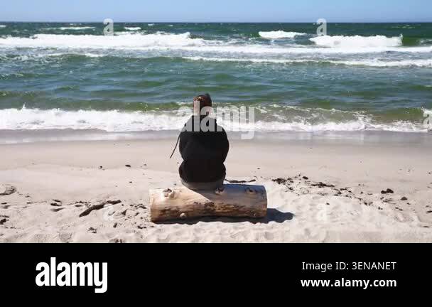 Woman dressed in black coat sits quietly on driftwood log facing gentle ...
