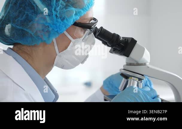 Female scientist wearing a mask and glasses, using a microscope in a ...