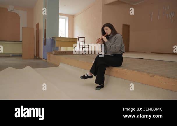 Talented young woman sits on a wooden stage in an empty, pastel-colored ...