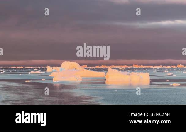 Icebergs float in a bay during sunset. The sun sets behind mountains ...