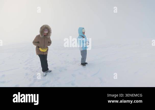 Side view of two children walking by hands down snowy slope in foggy ...