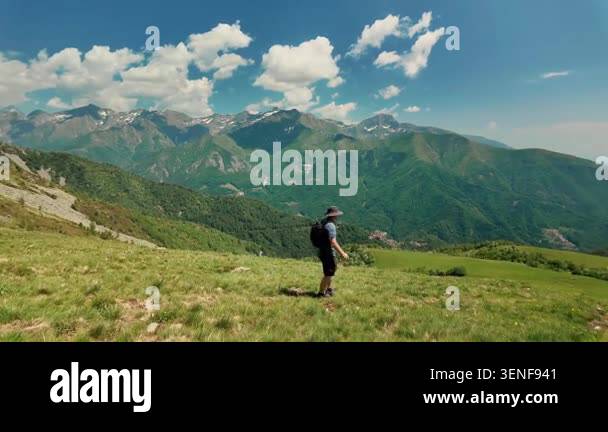 Hiker walking on green alpine slope waving hand toward camera in ...