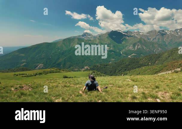 Hiker sitting on green alpine slope enjoying sweeping view of dramatic ...
