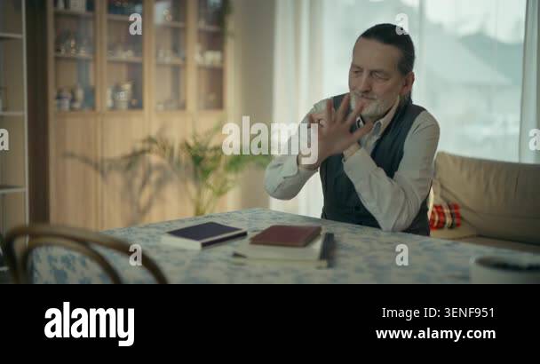 Older man seated at a table in a cozy living room, gesturing while ...