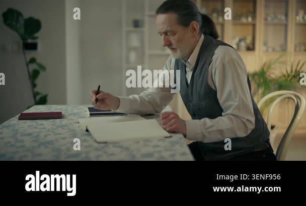Side view of an older bearded man with long hair writing in a notebook ...