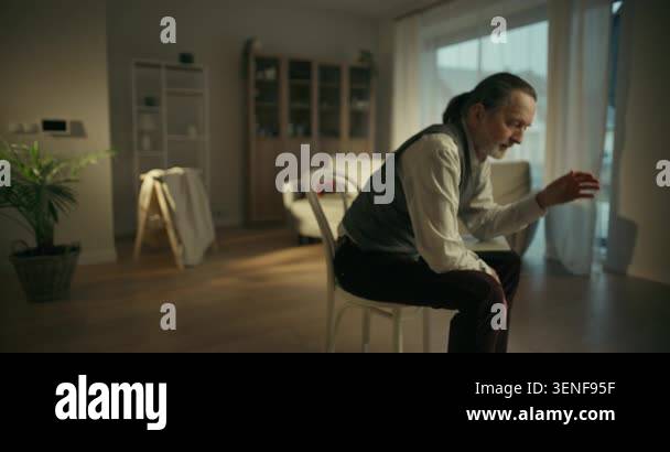 An elderly man sits in profile on a chair in a warm living room ...