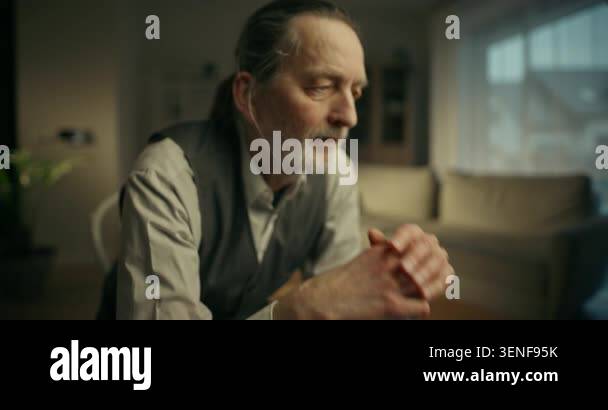 An elderly man with gray hair and a beard sits by a window in a softly ...