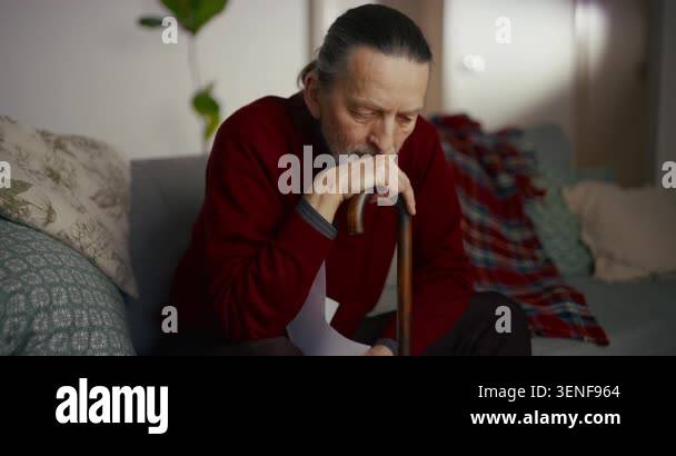 An elderly man in a red sweater sits on a sofa at home, leaning forward ...