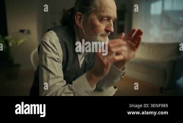 An elderly man sits indoors near a window in side profile, raising his ...