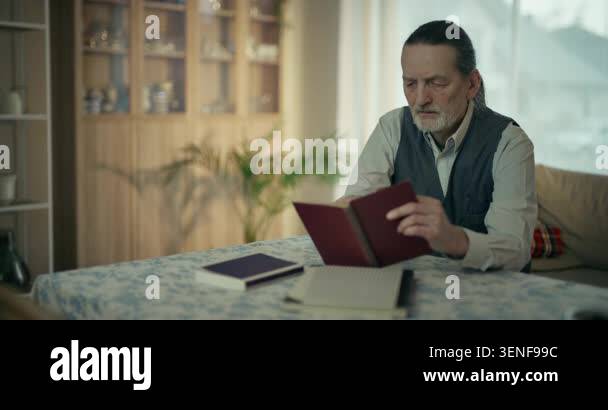 An older man sits at a table in a bright living room, reading an open ...