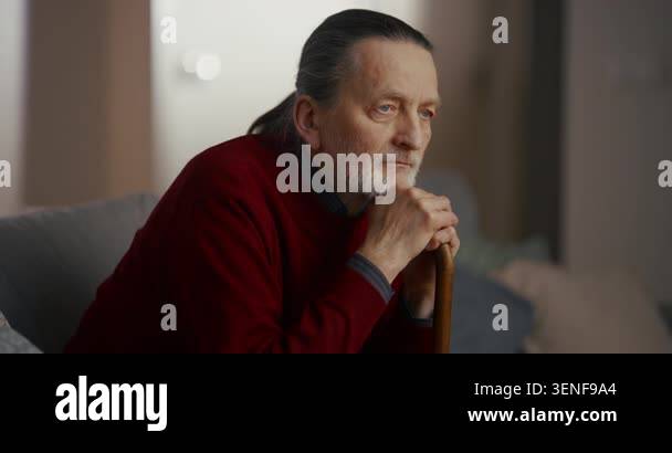 An elderly man with gray hair and a beard sits indoors by a window ...