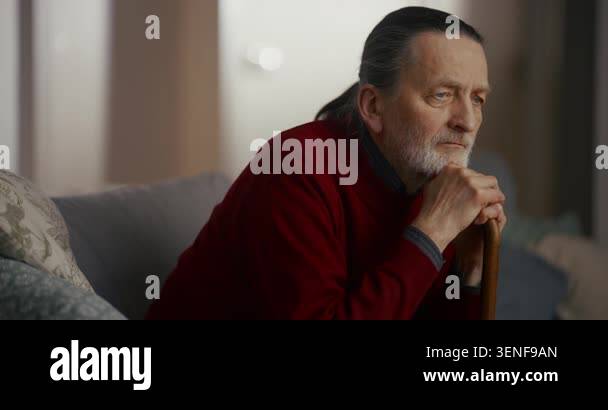 An elderly man in a red sweater sits indoors beside a bright window ...