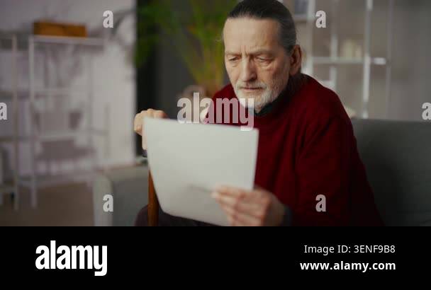 Senior man with gray hair and beard sits indoors, looking down at a ...