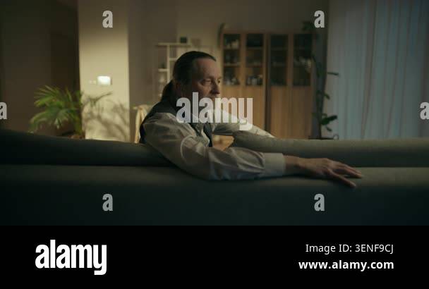 Middle-aged man sits in side profile on a sofa in a warm, softly lit ...