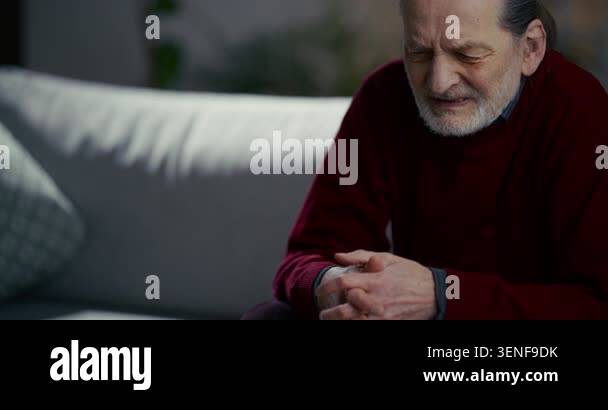 An elderly man sits alone on a sofa indoors with clasped hands and a ...