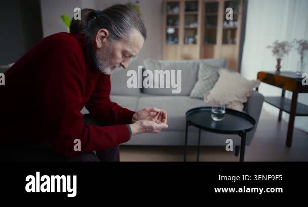 Older man seated in a home living room, looking down at his hands with ...