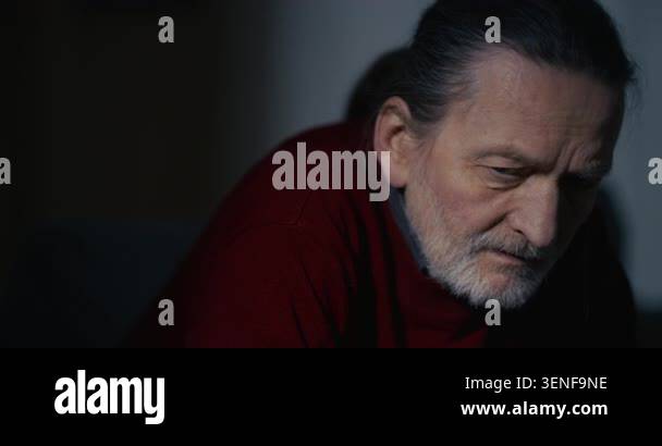 Close-up of an older man in a red shirt looking downward with a pensive ...