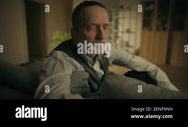 An elderly man with a gray beard sits back on a sofa with eyes closed ...