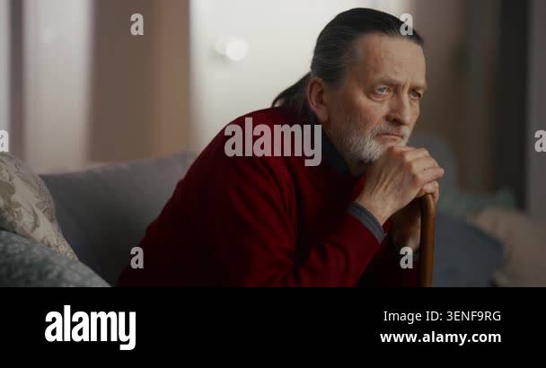 Elderly man in a red sweater sits on a sofa indoors, leaning forward ...