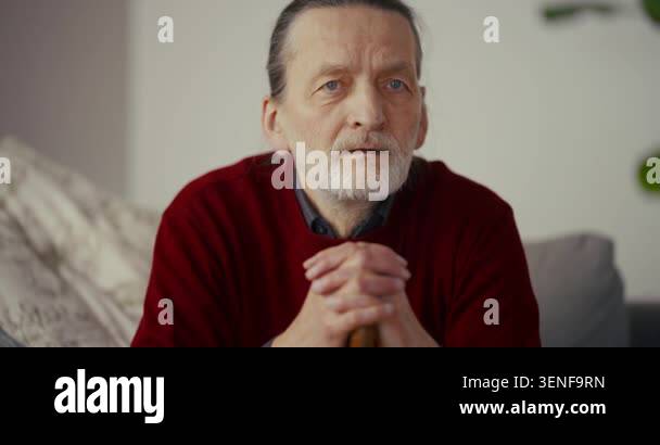 An elderly man with gray hair and a beard sits on a couch indoors ...