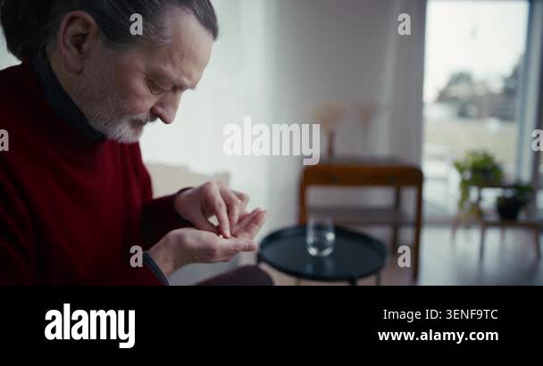 Senior man sits by a sunlit window at home, looking down at his hands ...