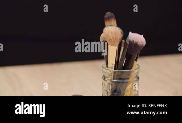 Several makeup brushes stand upright in a clear glass jar on a light ...