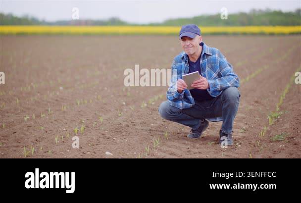A man crouches beside neat crop rows in a cultivated field while using ...
