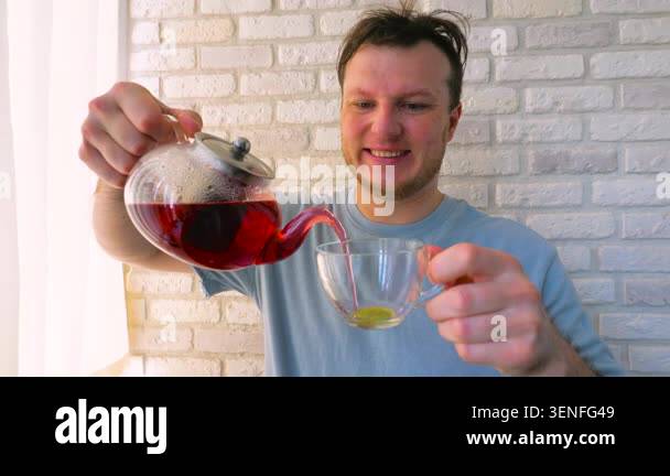 Pour red tea into a cup from a transparent teapot. A young man drinks ...