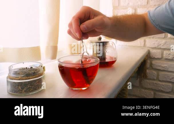 A young man stirs tea in a transparent cup with a spoon. Drink red tea ...