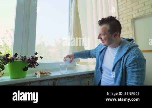 A young man pours yogurt into a glass while sitting near the window in ...