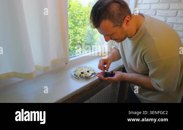 A man sprinkles blue berries into cottage cheese in the kitchen ...