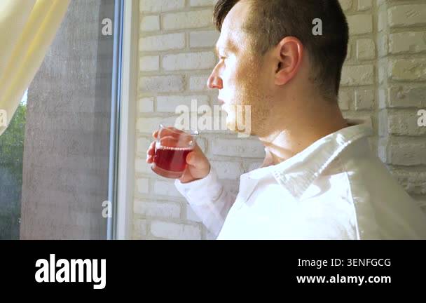A young man drinks red juice against the background of a window. A man ...