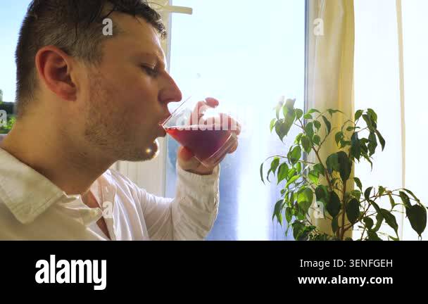 A young man drinks red juice against the background of a window. A man ...