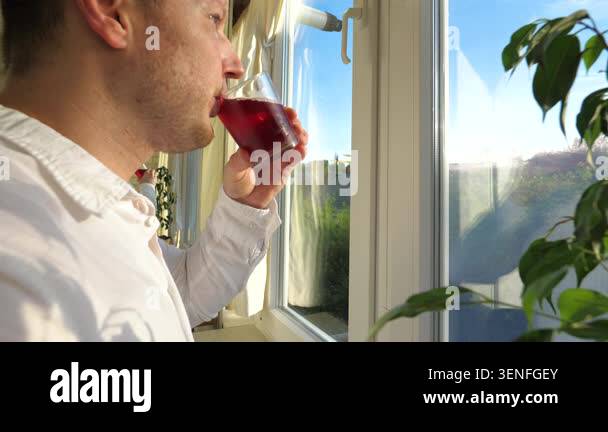 A young man drinks red juice while looking out the window. A man drinks ...