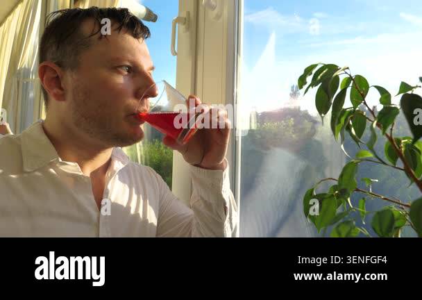 A young man drinks red juice while looking out the window. A man drinks ...
