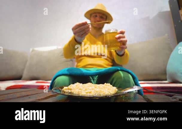 Eating porridge with honey at home at a wooden table. A young man pours ...