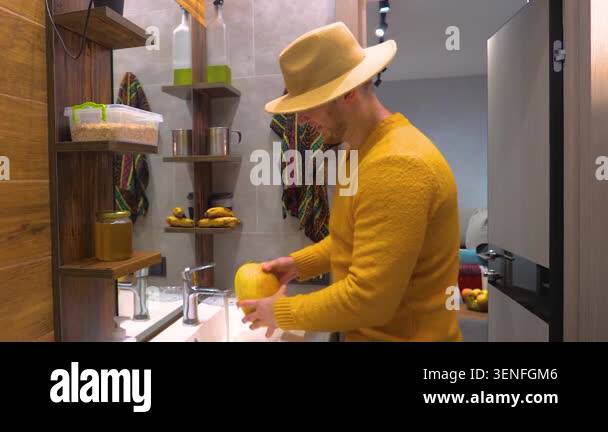 A young man washes a melon fruit under a water tap in an apartment. A ...