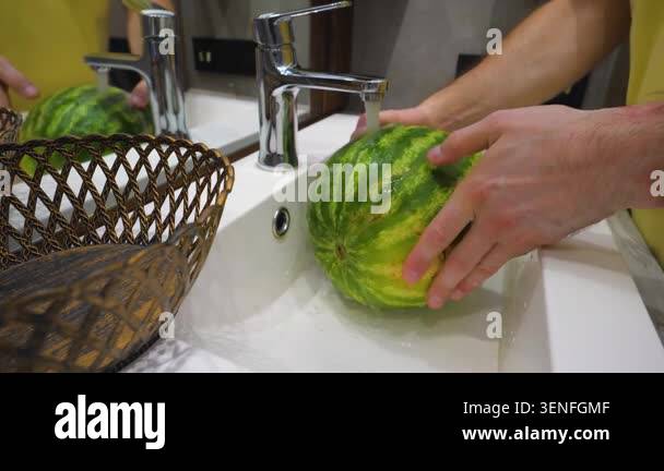 A young man washes a watermelon under the water tap in the kitchen ...